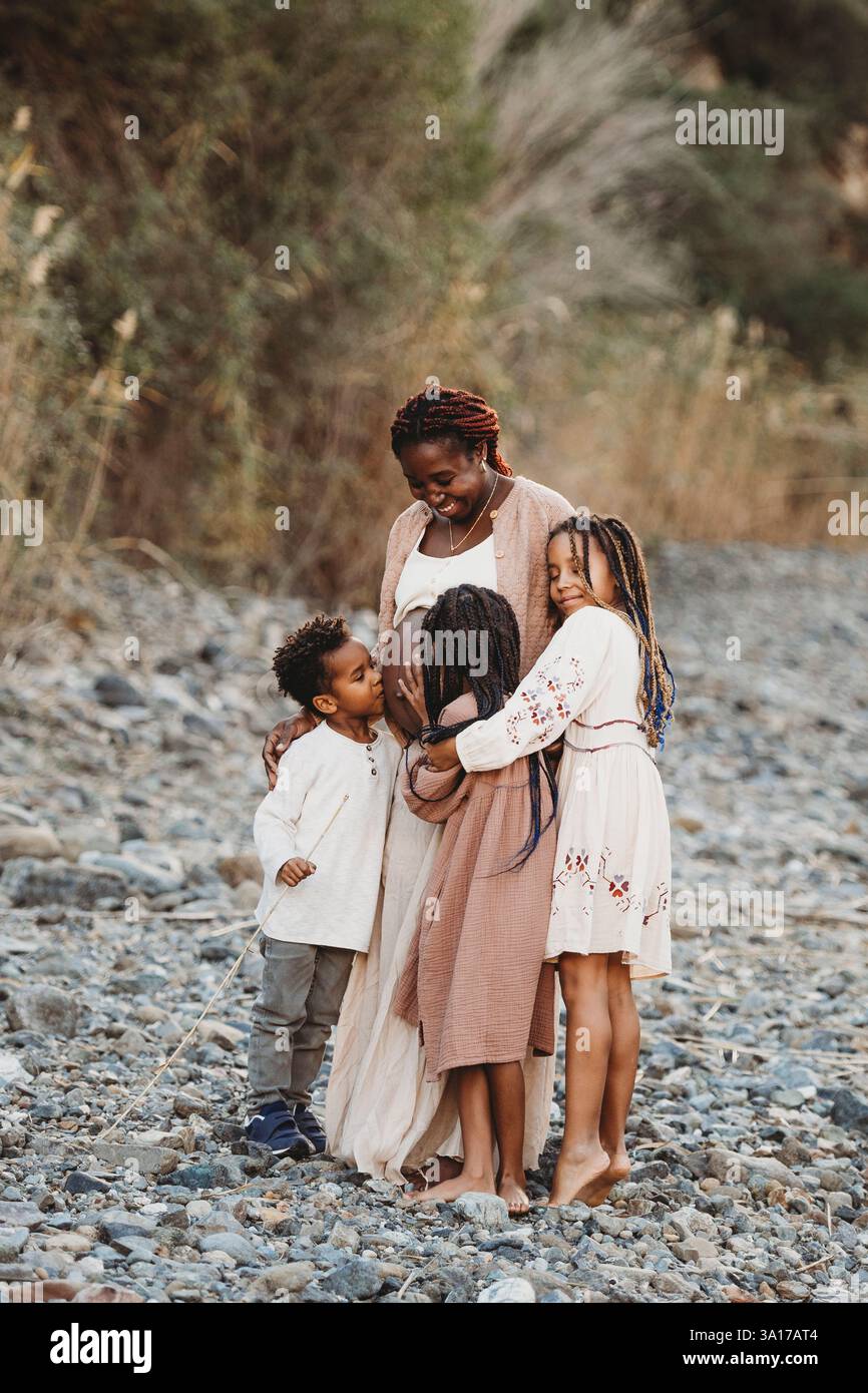 Smiling black pregnant mom mix race children kissing belly on rocks ...