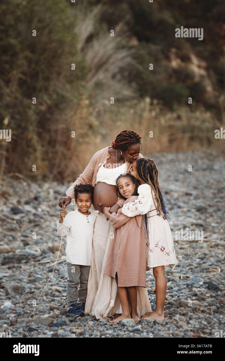 Smiling black pregnant mom mix race children kissing belly on rocks ...