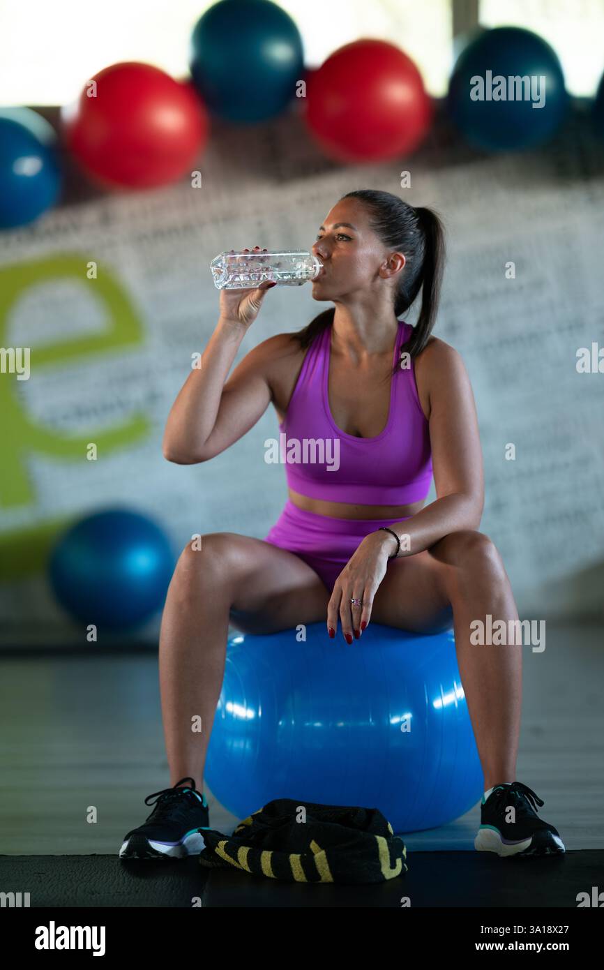 A fit woman takes a well deserved break in the gym, sipping water from her bottle after an ...