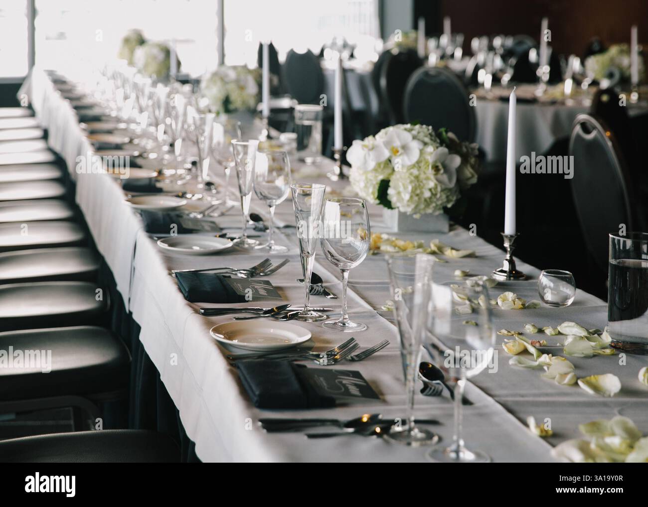 A long banquet table is set for a formal gathering, featuring elegant glassware and floral ...