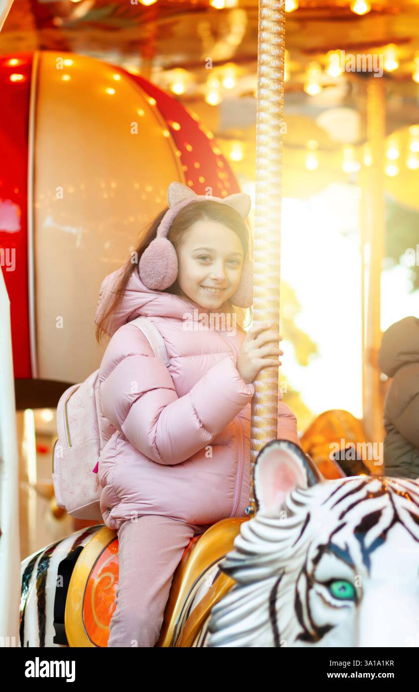 Happy girl wearing pink earmuffs joyfully riding a carousel tiger at an ...