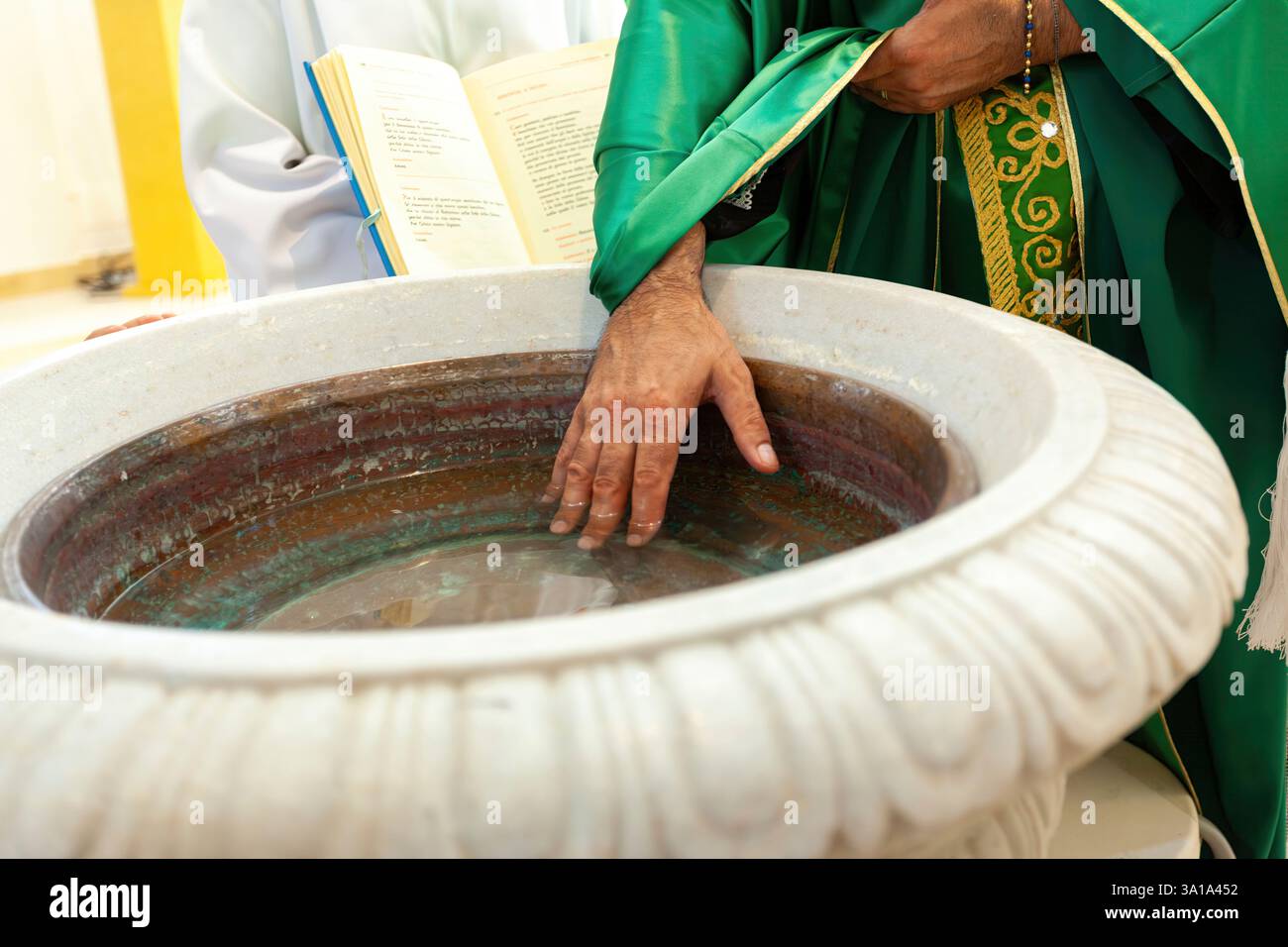 Priest wearing green vestments blessing the water of a marble baptismal font with his hand ...