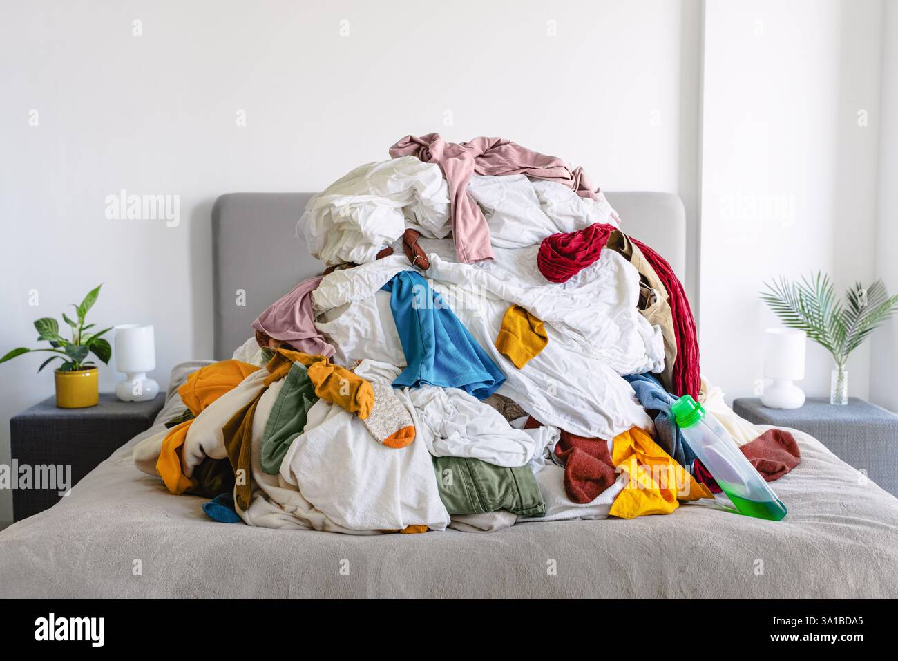 A large pile of mixed laundry covers a bed, with a detergent bottle ...