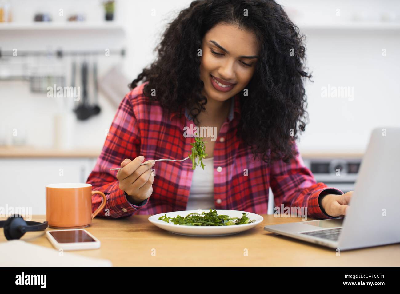 African American young woman in kitchen eating salad, working on laptop ...