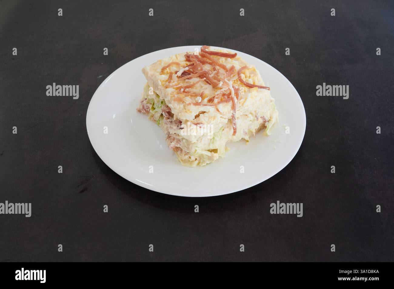 A slice of food sits on a white plate. The food is a layered dish with ...