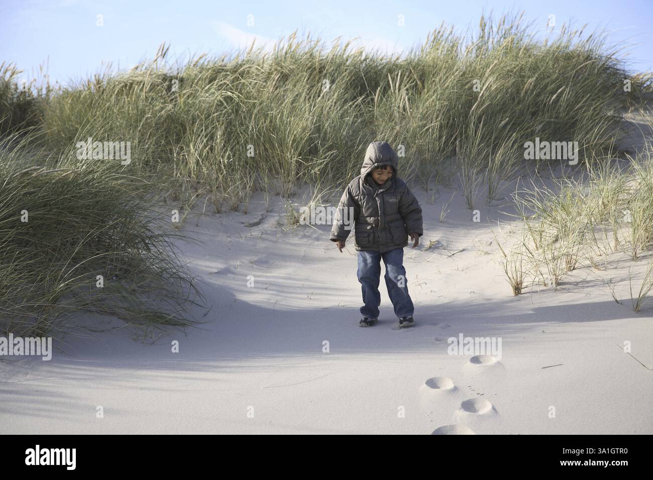 Boy wearing jacket watching his foot prints on sand, Skagen, Denmark ...