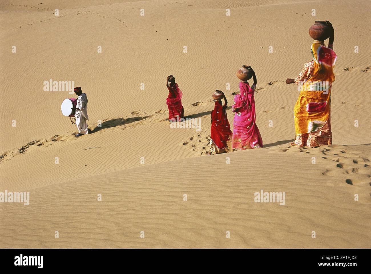 Rajasthani Women carrying water pots and man playing drum walking in sand, Khuhri, Jaisalmer ...
