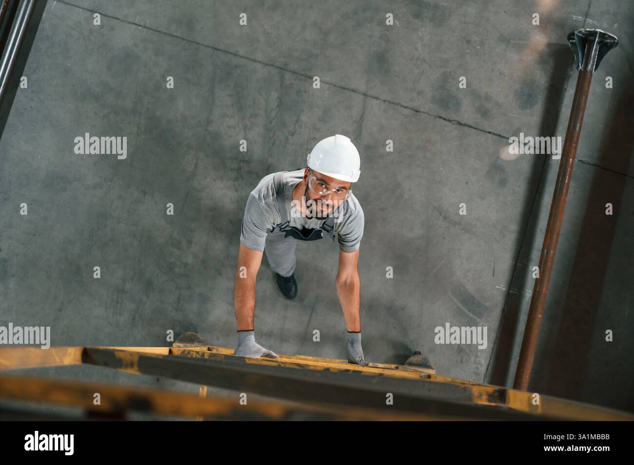 Going up on the ladder, top view. Young factory worker in grey uniform ...