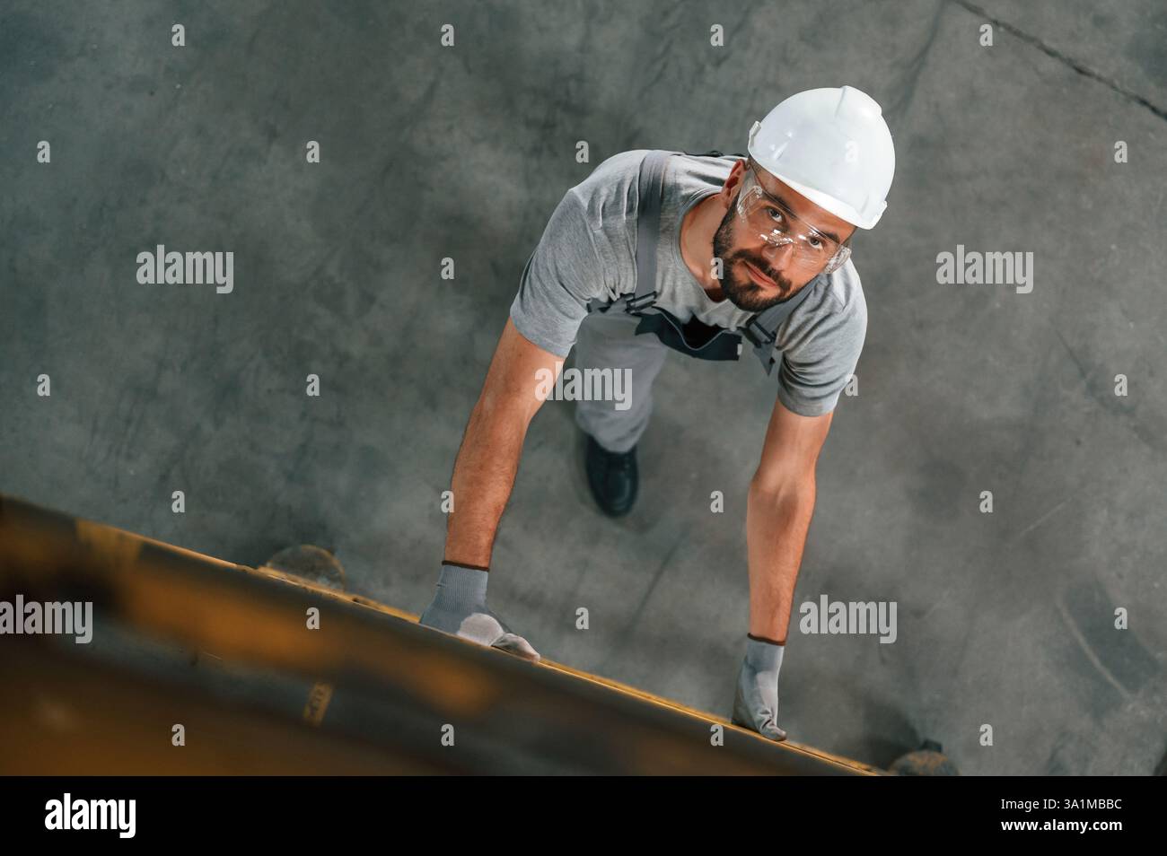 Going up on the ladder, top view. Young factory worker in grey uniform ...