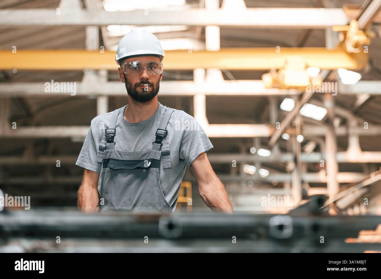 In glasses and white hard hat. Young factory worker in grey uniform ...