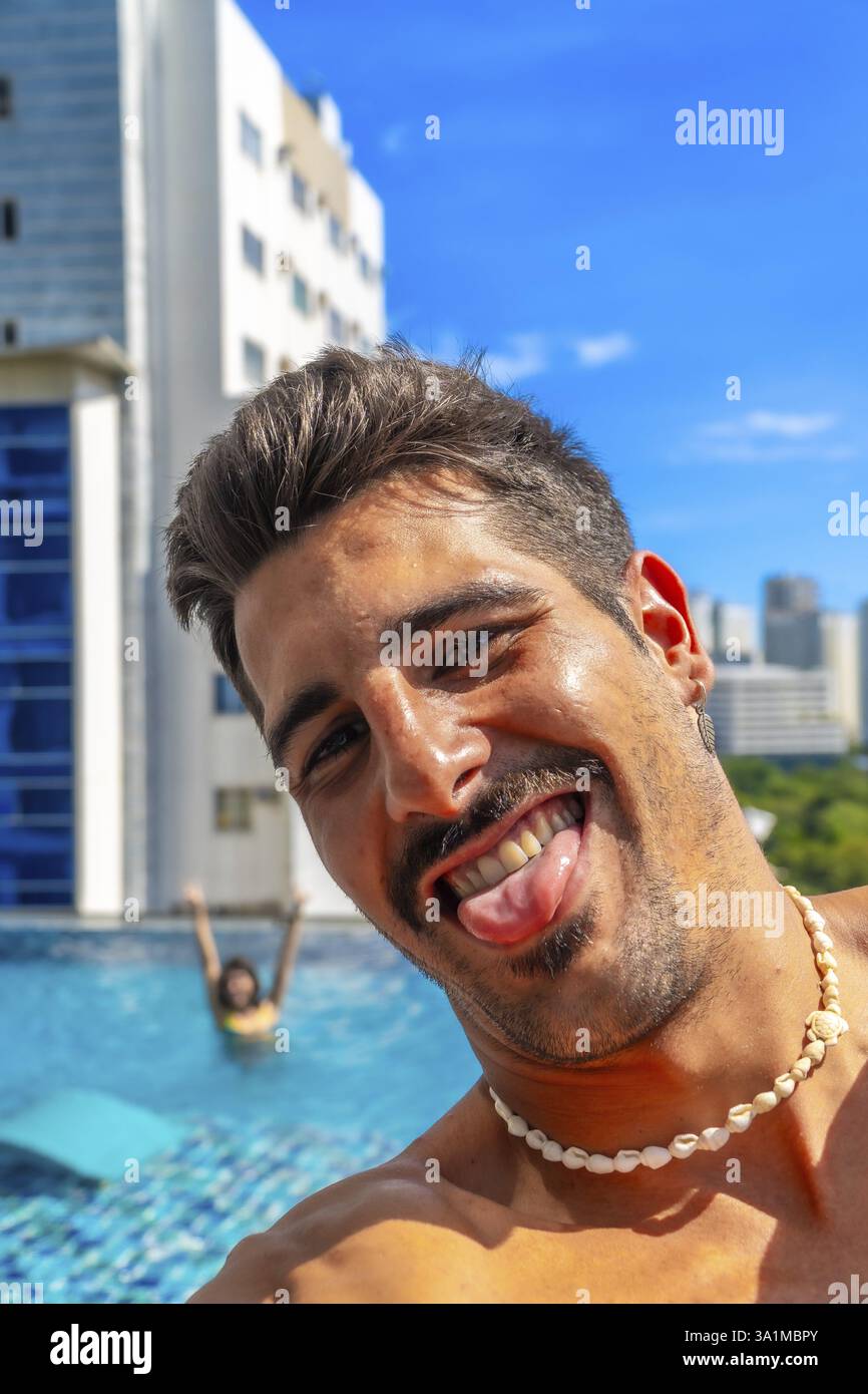 Young man wearing shell necklace sticking out his tongue by hotel rooftop pool in cebu city ...