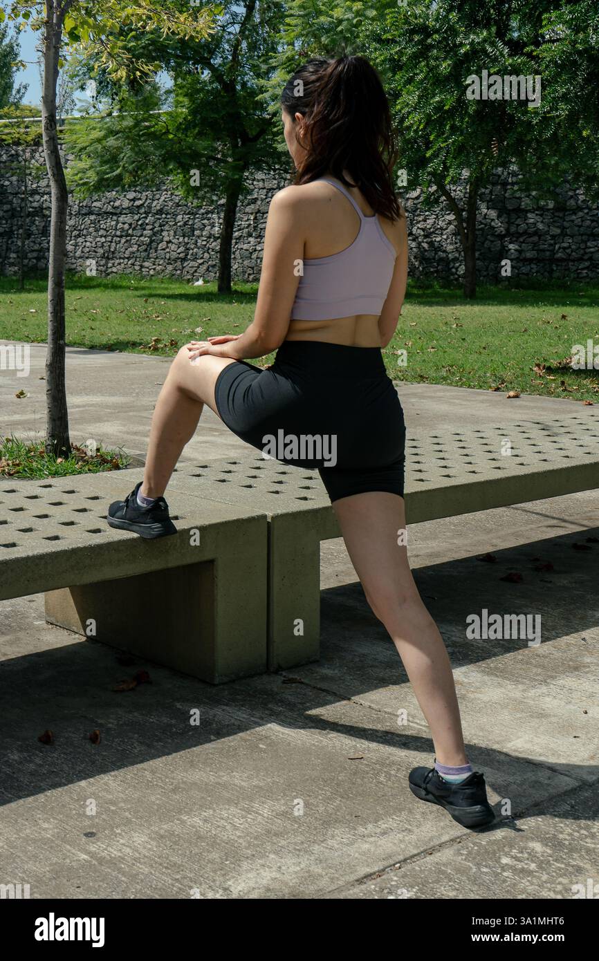 Fit woman performing leg stretches on a bench in a green park ...