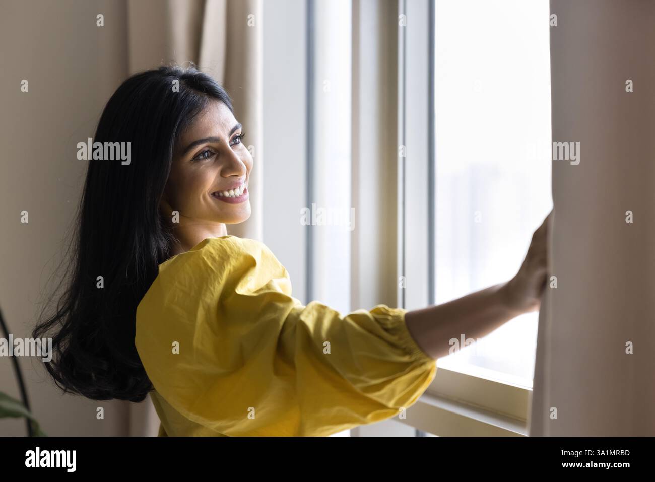 Young Indian woman standing by window, pulling curtains with smile ...