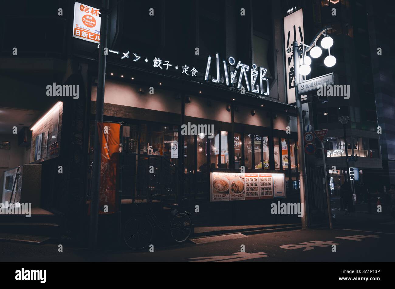 A warmly lit ramen shop stands quietly amidst the city’s nightscape ...