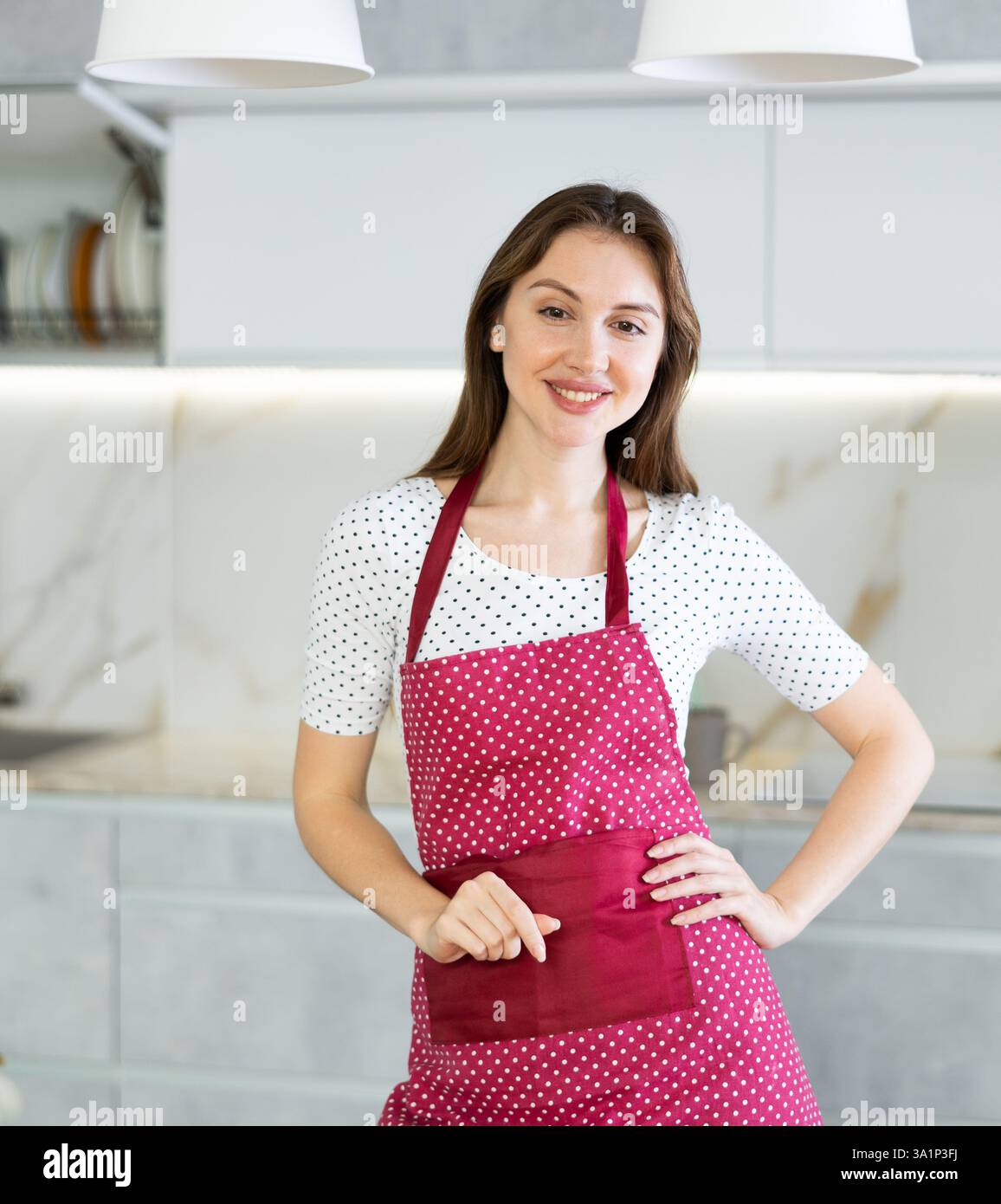 Girl in apron stands in kitchen and put one hand on belt and ...