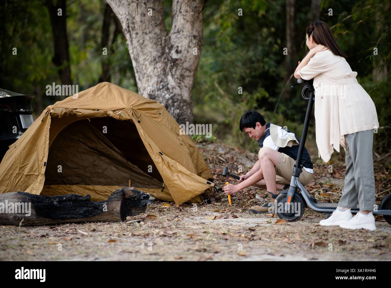 The woman is holding the scooter while watching the man setting up their camping tent. Family ...