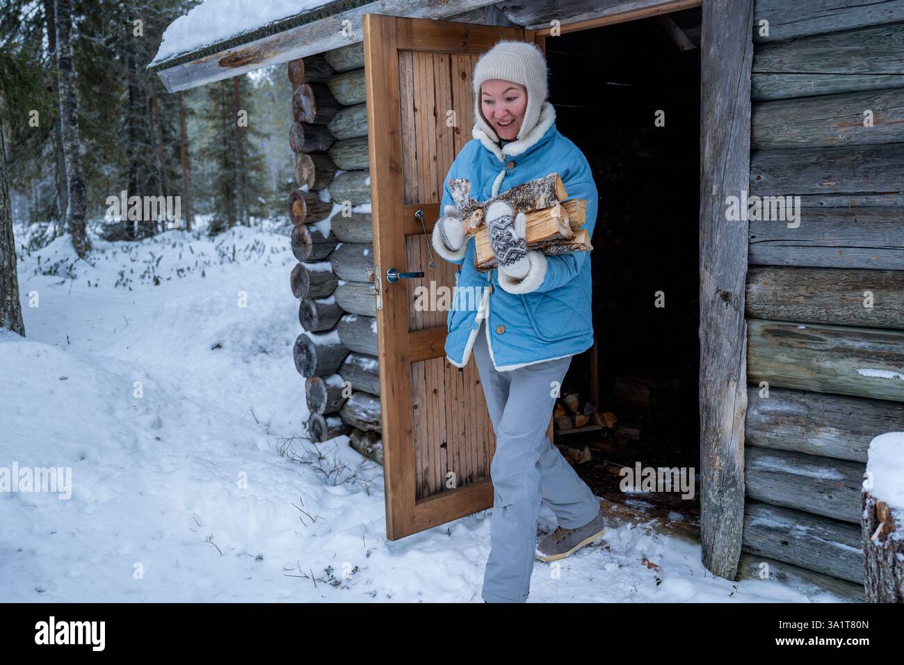 Woman in blue winter coat carrying firewood outside a rustic log cabin ...