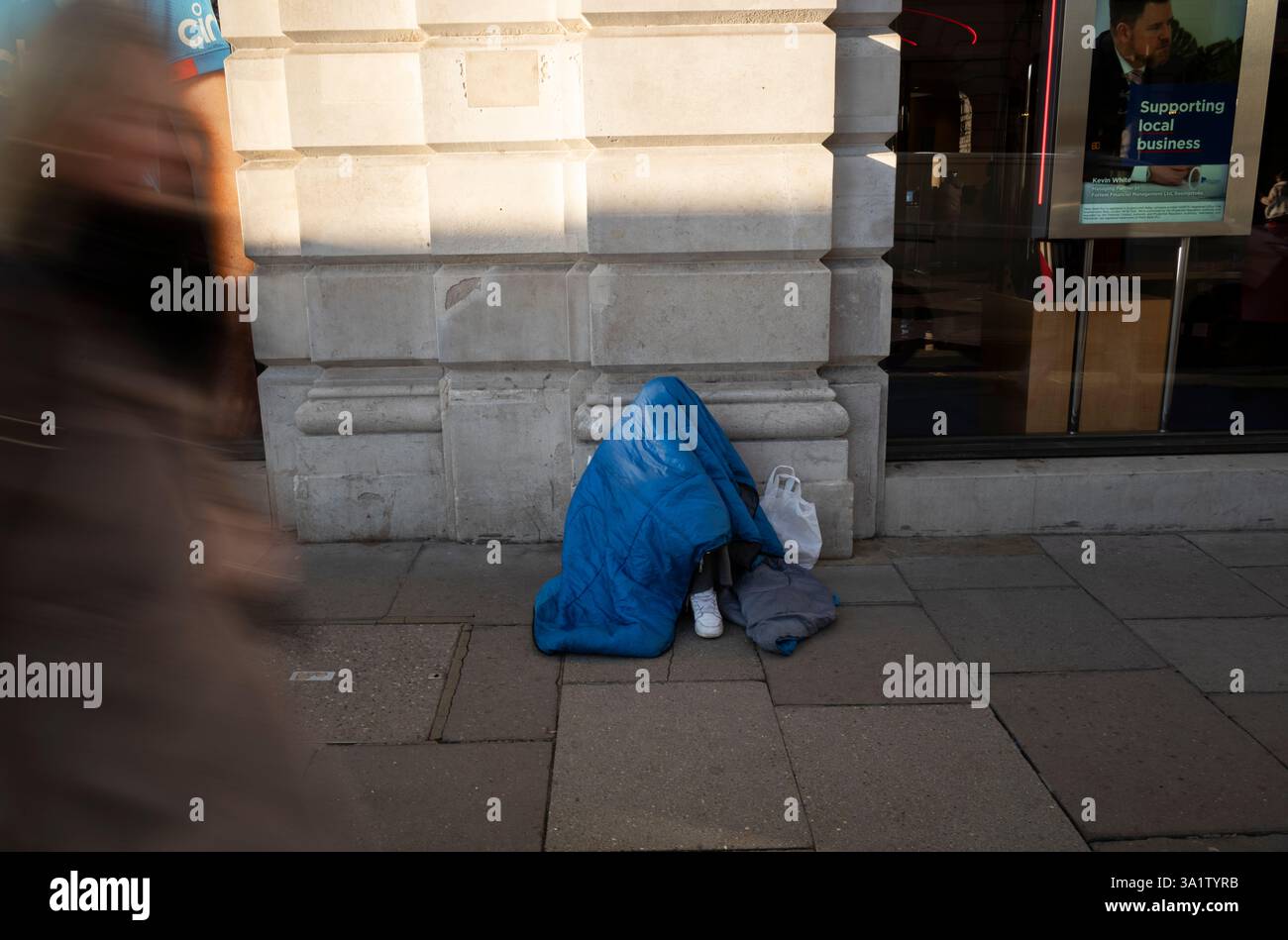 Homeless woman covered by a sleeping bag whilst sat on the pavement ...