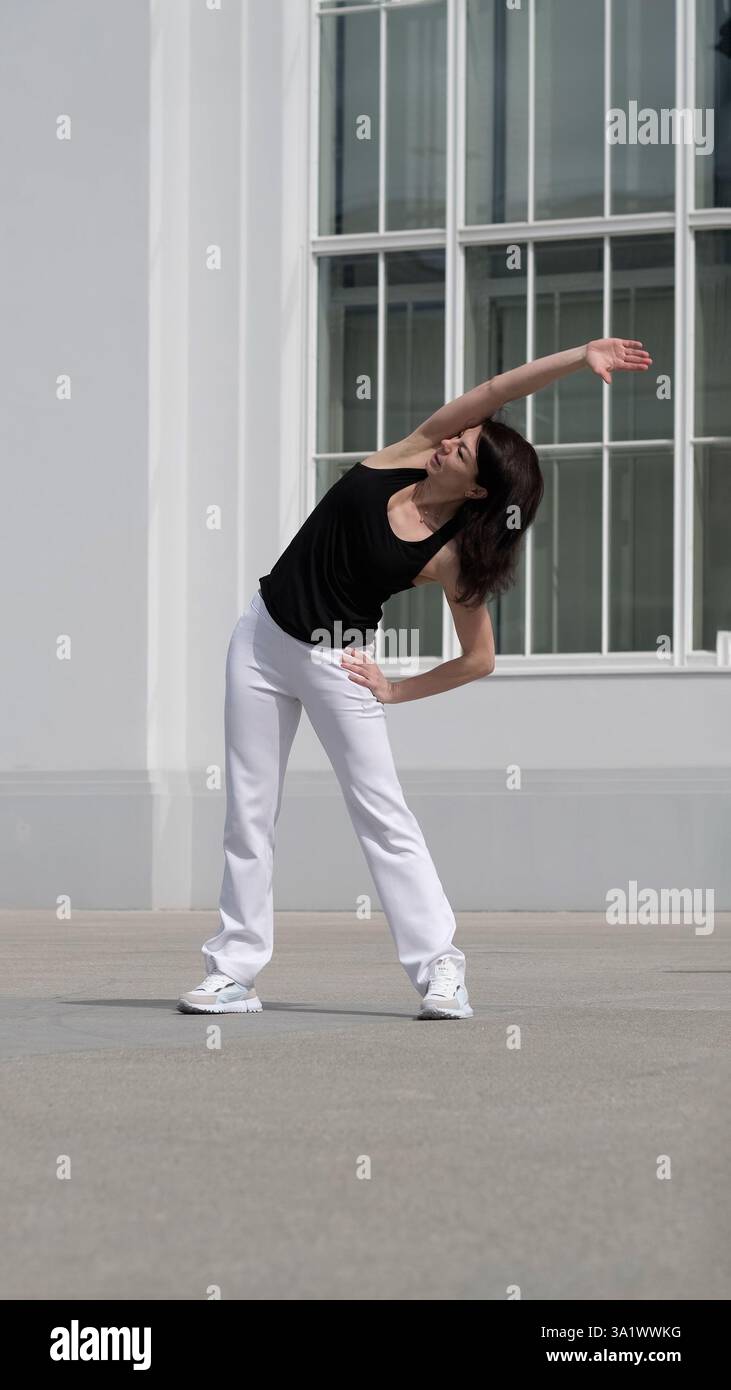 A woman in a black top and white pants stretches gracefully against a modern building. The scene ...