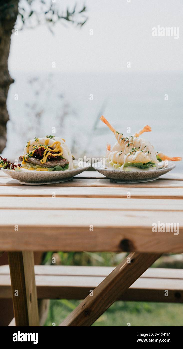 Two plates of food sit on a wooden table by the ocean. The food is a ...