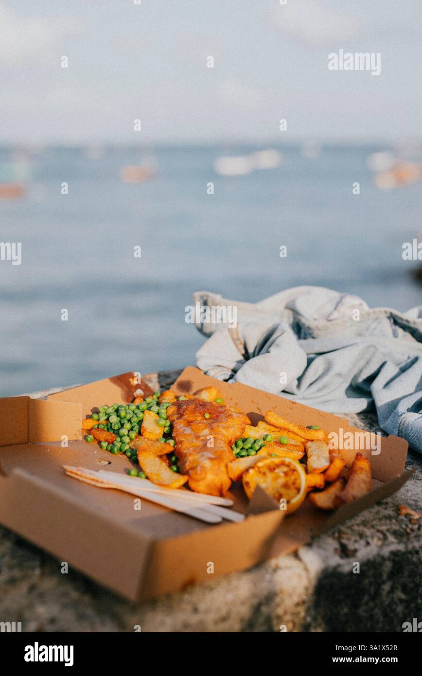 A box of fish and chips is sitting on a ledge near the water. The fish ...