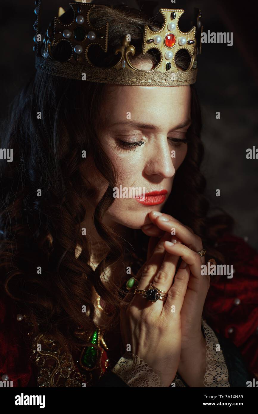 medieval queen in red dress with crown praying on dark gray background ...