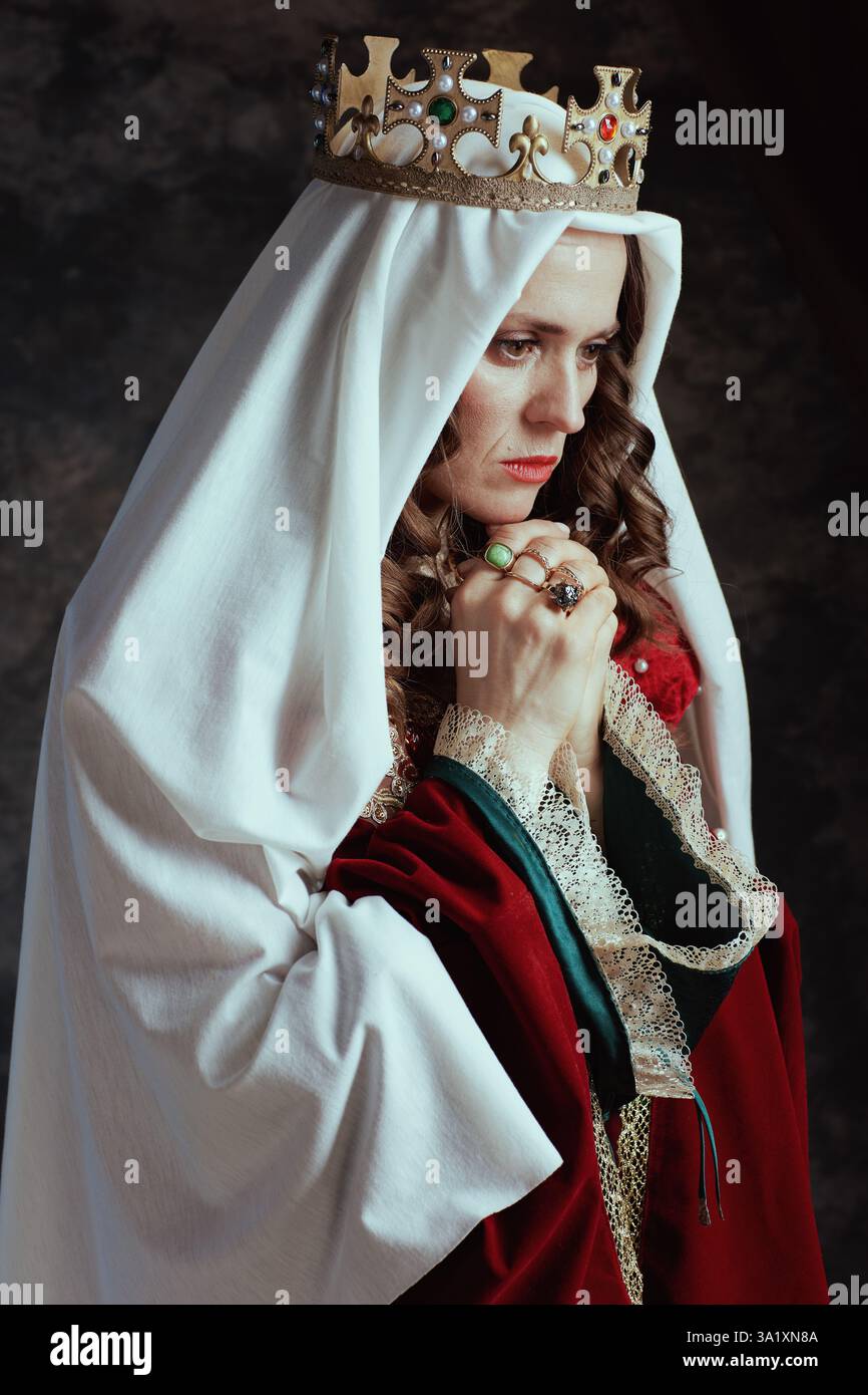 medieval queen in red dress with veil and crown praying on dark gray ...