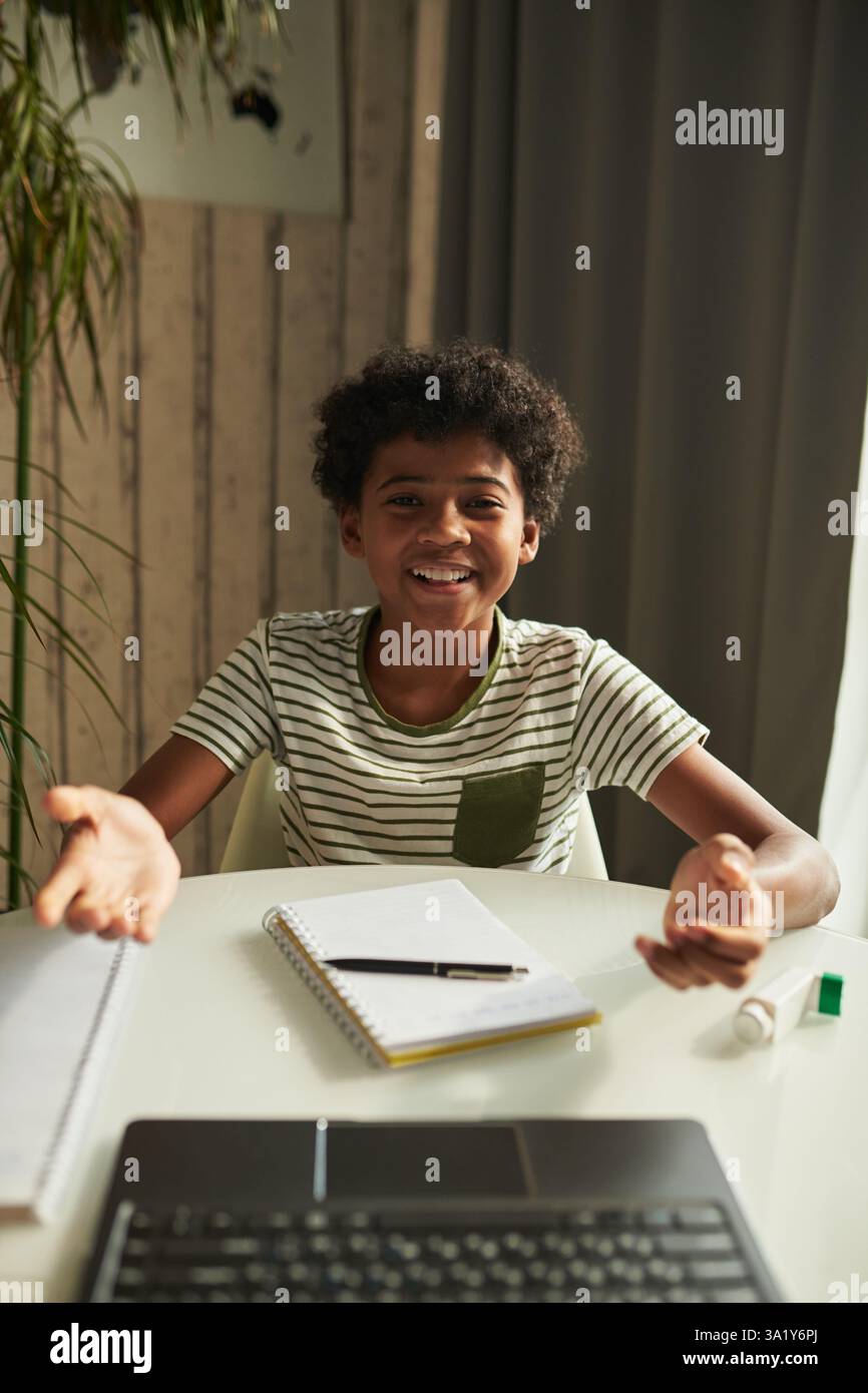 Happy schoolboy sitting by table in front of laptop screen and ...