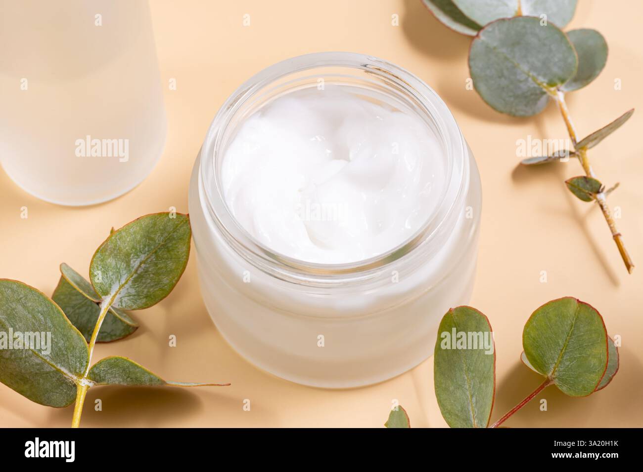 open glass gar with face cream and eucalyptus against beige background ...