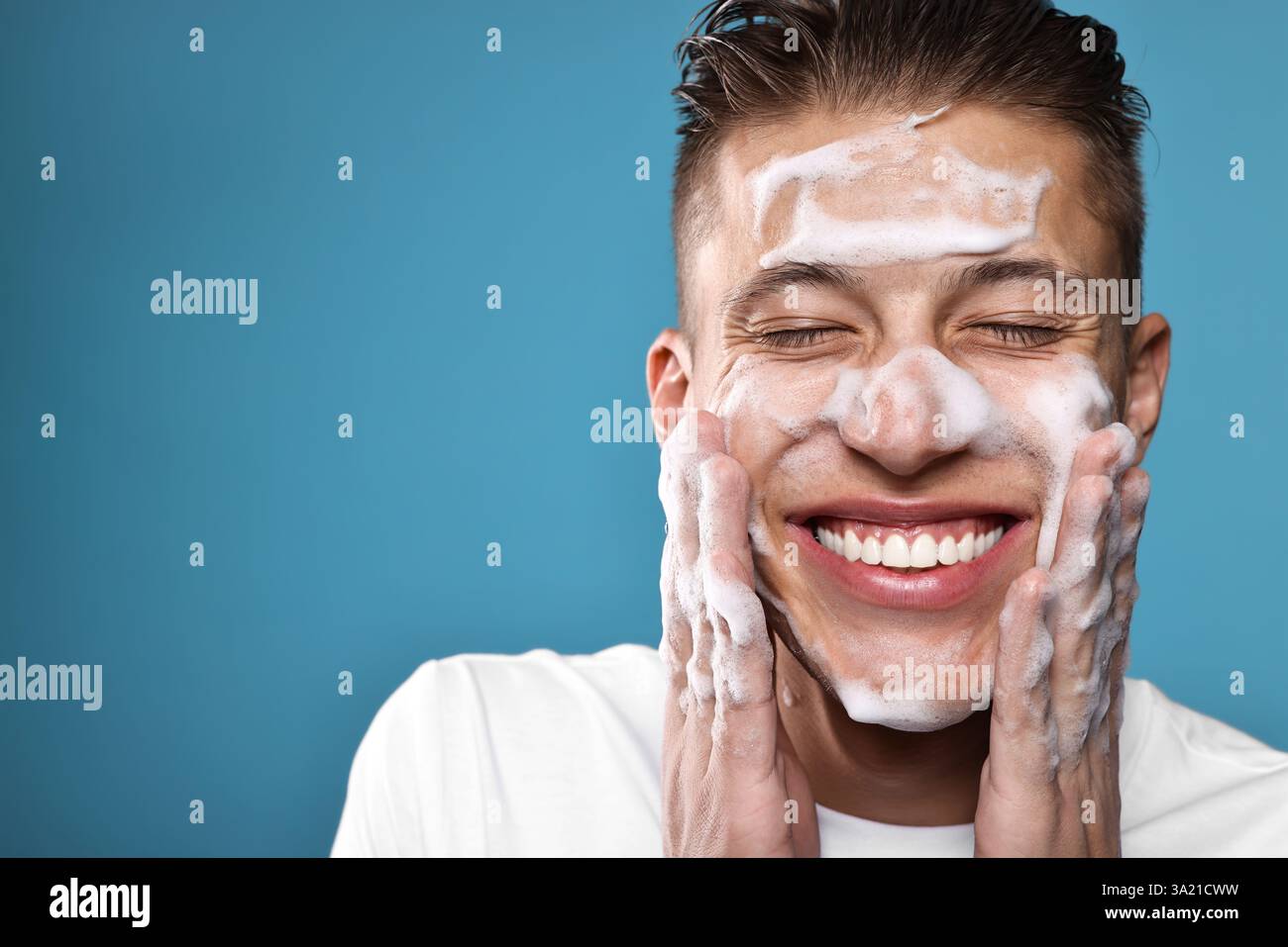 Smiling man washing his face with cleansing foam on blue background, closeup. Space for text ...