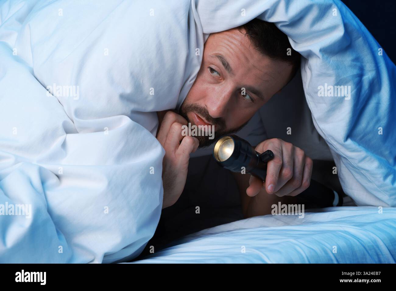 Fear of darkness. Scared man with blanket and flashlight on bed in room ...