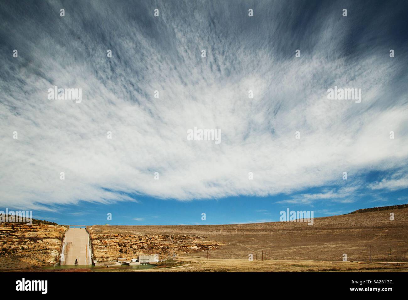 A dam wall and sluice overflow into a lake, with a blue sky and ...