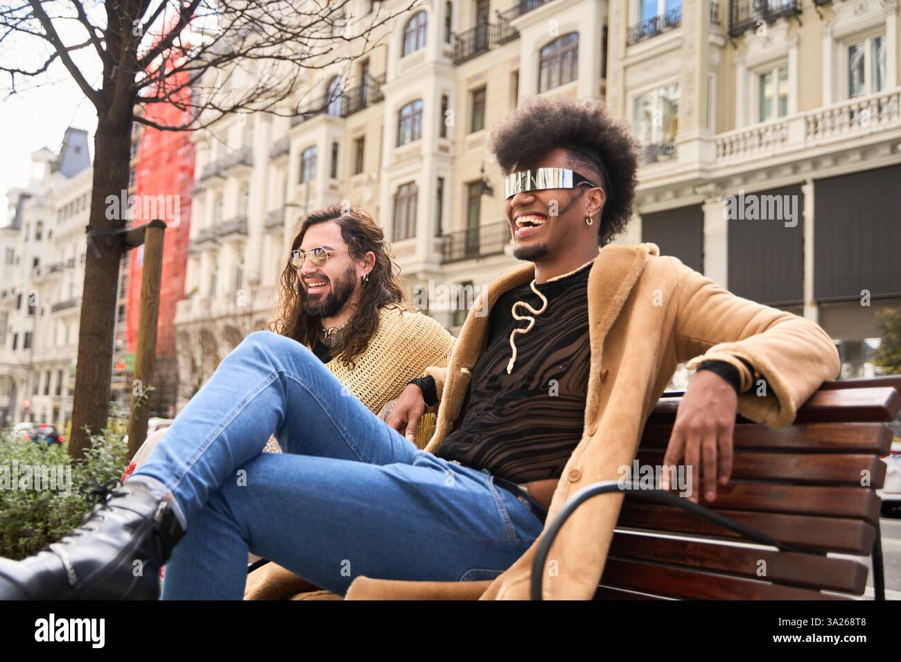 Two stylish non binary people wearing sunglasses are sitting on a bench ...