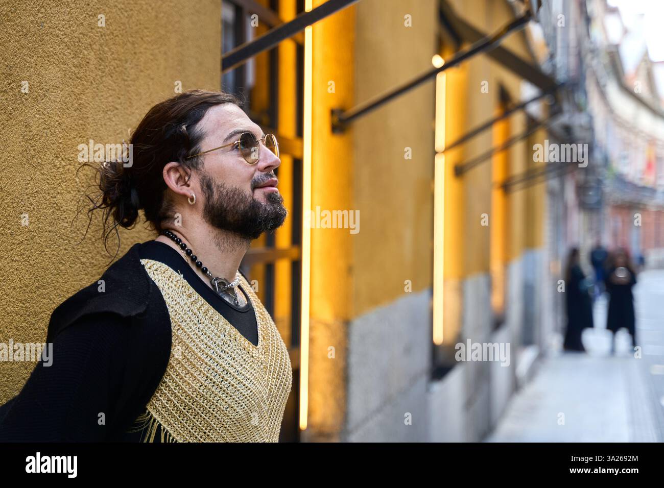 Stylish non binary person with long hair and beard looking away while posing in urban street ...