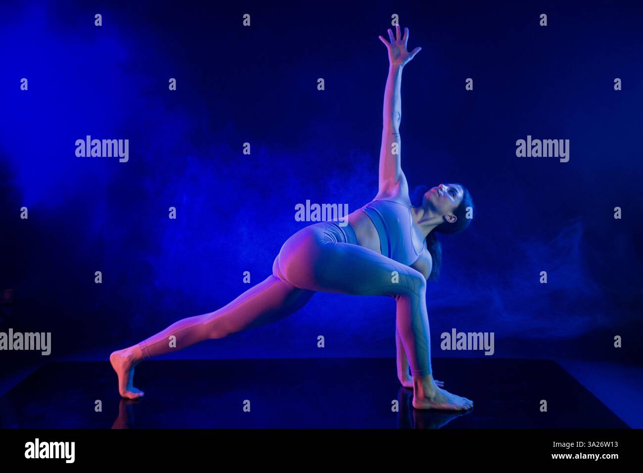 Woman in yoga pose with one arm extended, in a blue-lit studio with ...