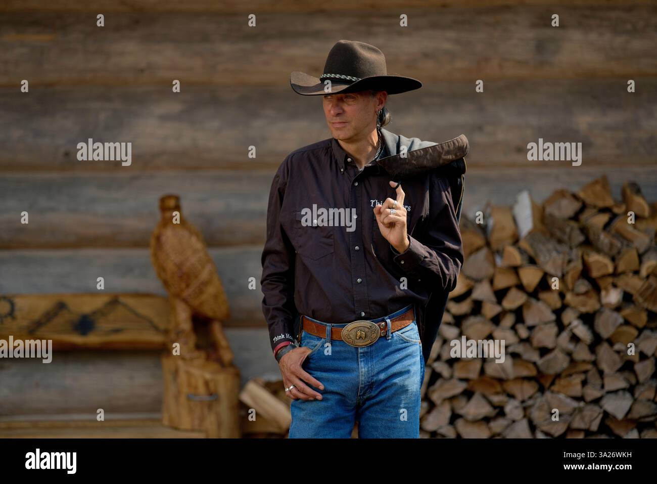Cowboy in a black shirt and hat stands by a log cabin with a stack of ...