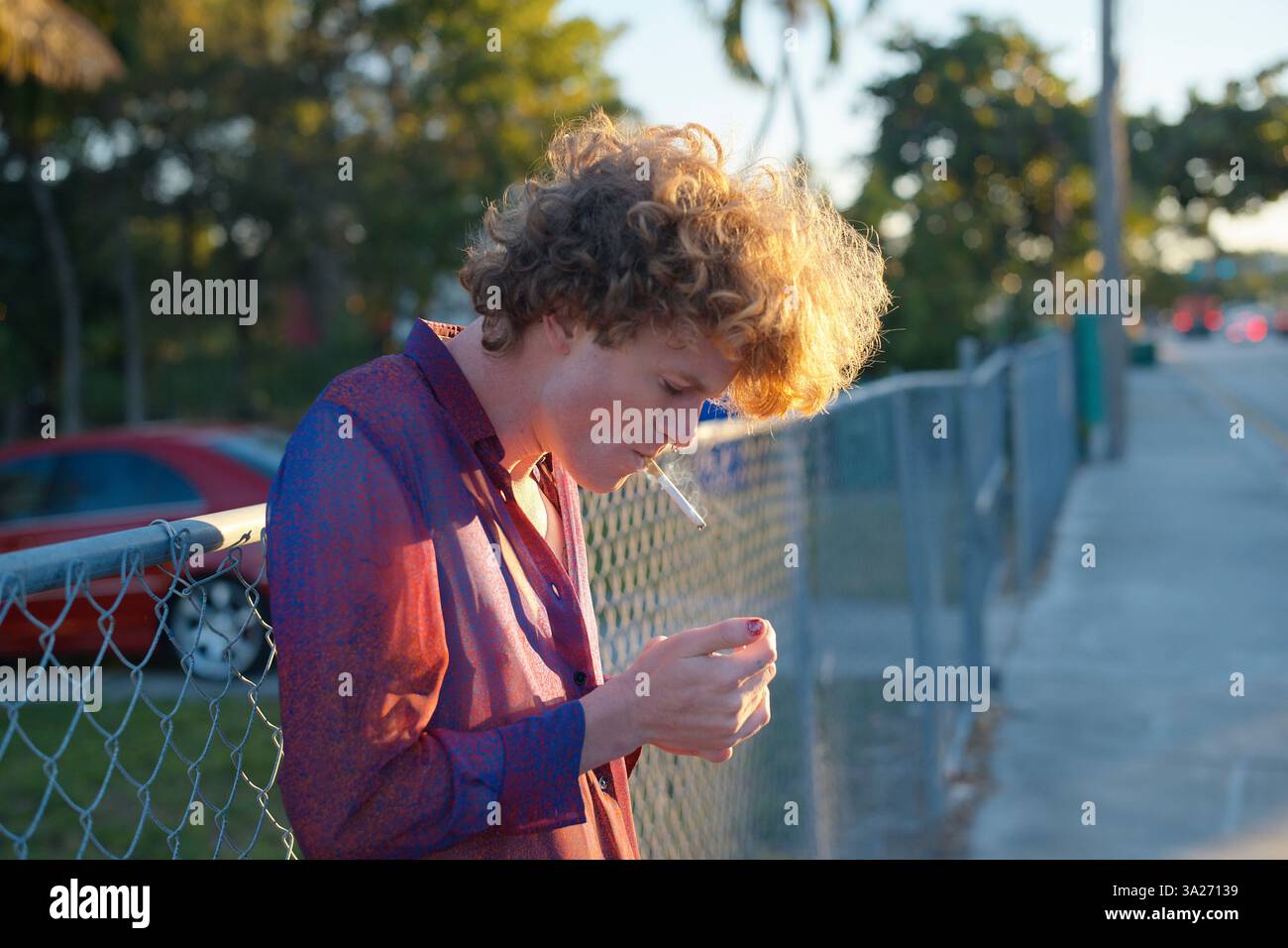 Curly-haired person lighting a cigarette by a chain-link fence on a ...