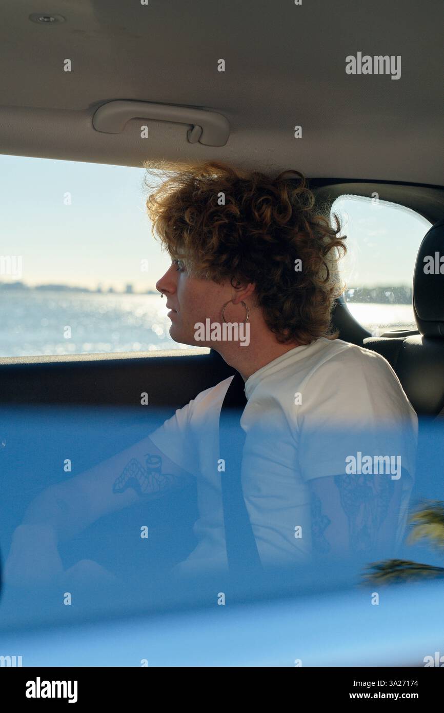 Young person with curly hair and tattoos looking out a car window at a ...