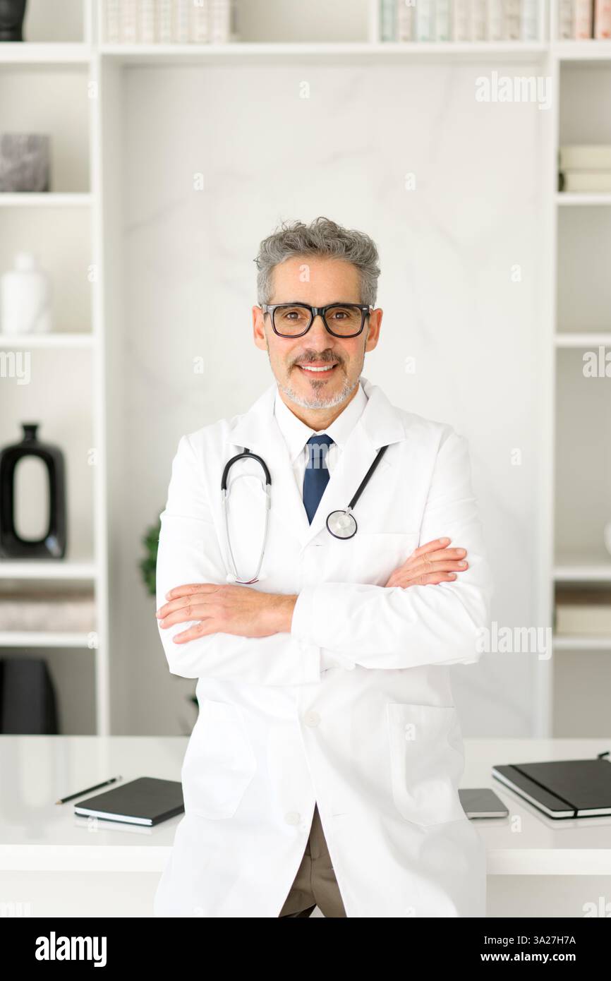 A mature doctor in a white coat with a stethoscope stands with hands ...
