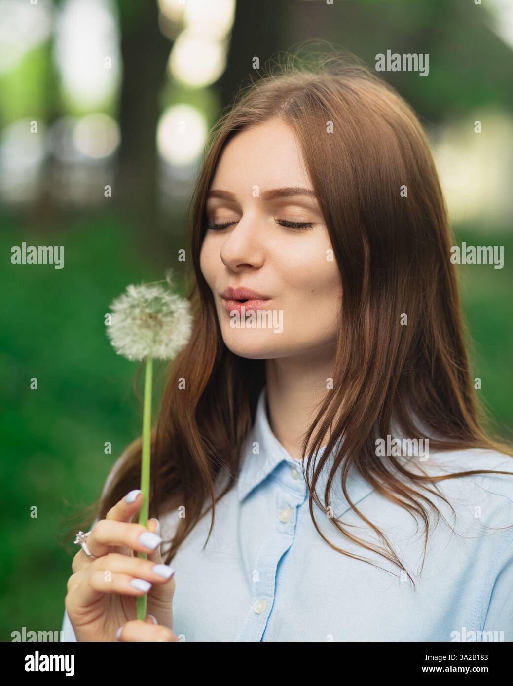 A beautifully serene young woman is gently blowing a dandelion in a ...