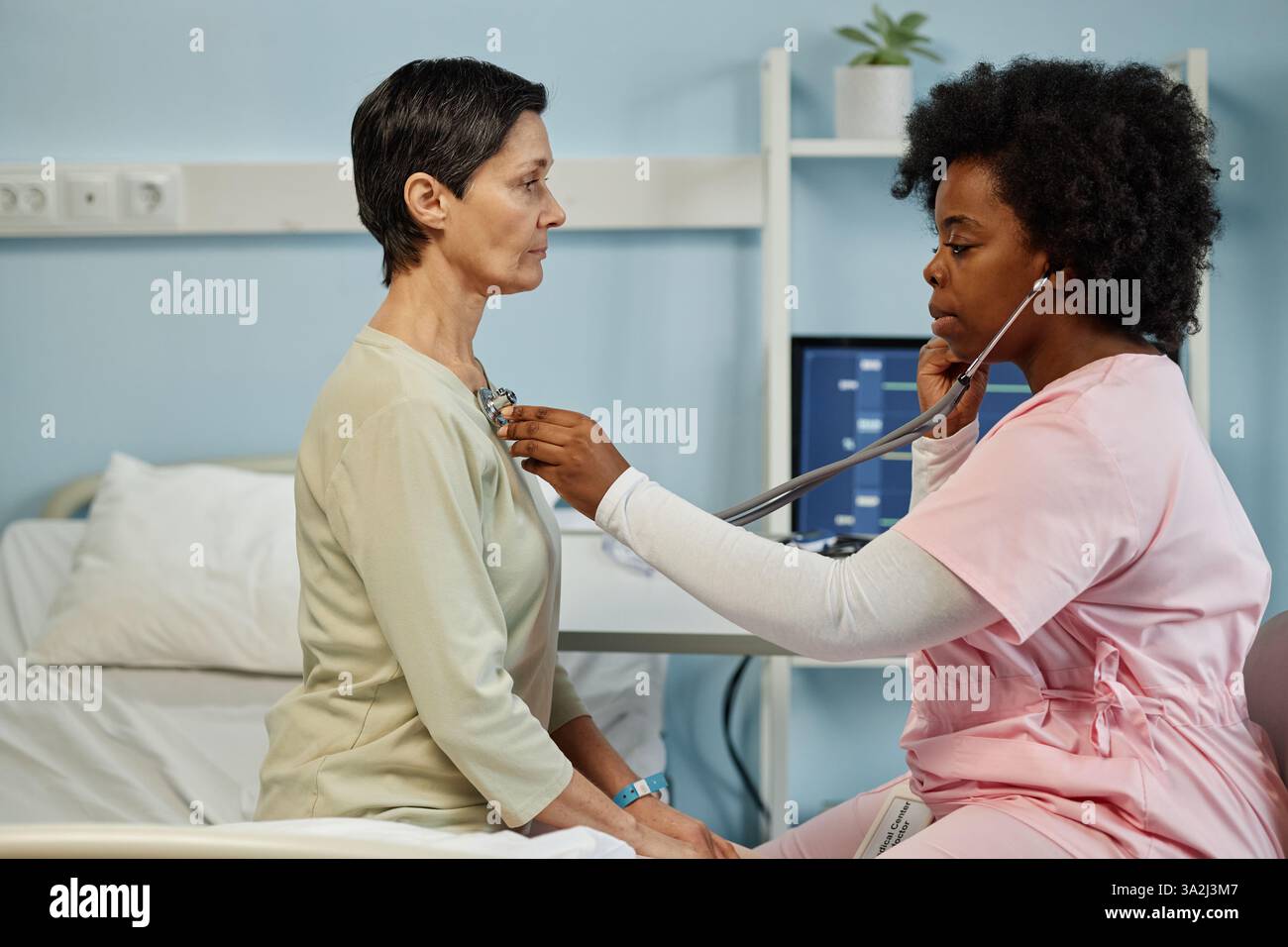 Nurse in pink uniform examining patient with stethoscope in clinic room ...