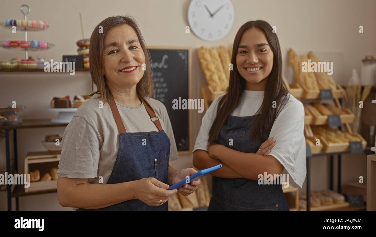 Two smiling female bakers in a bakery shop, one using a tablet and the ...