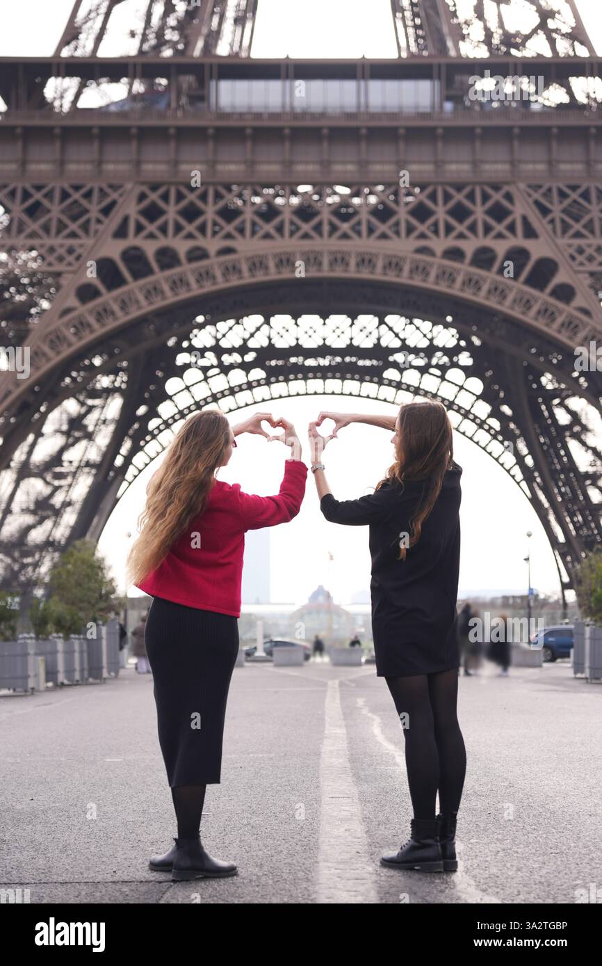 Two young women stand beneath the Eiffel Tower, forming a heart shape ...