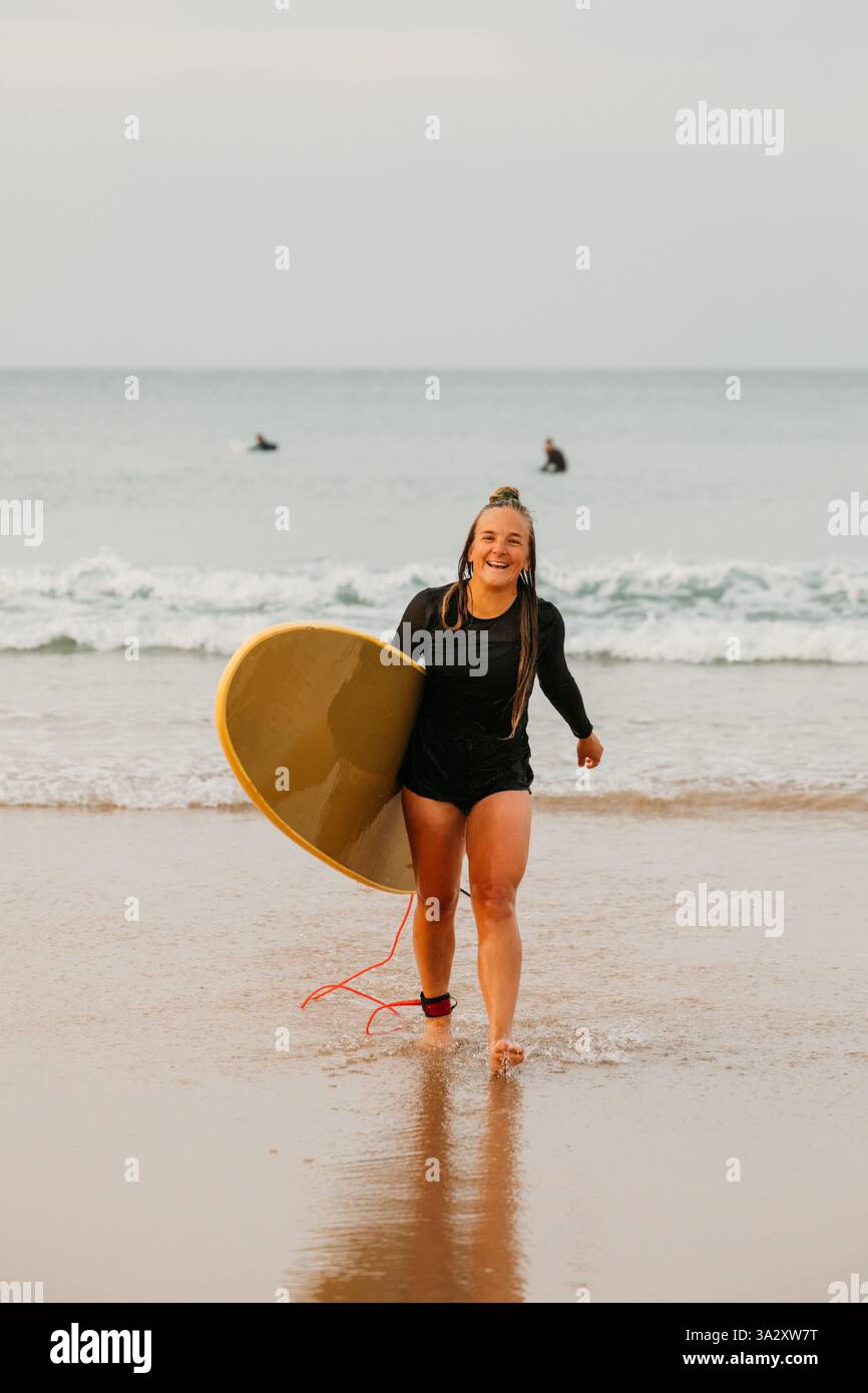 A happy female surfer walks back from the ocean, carrying her bo Stock ...