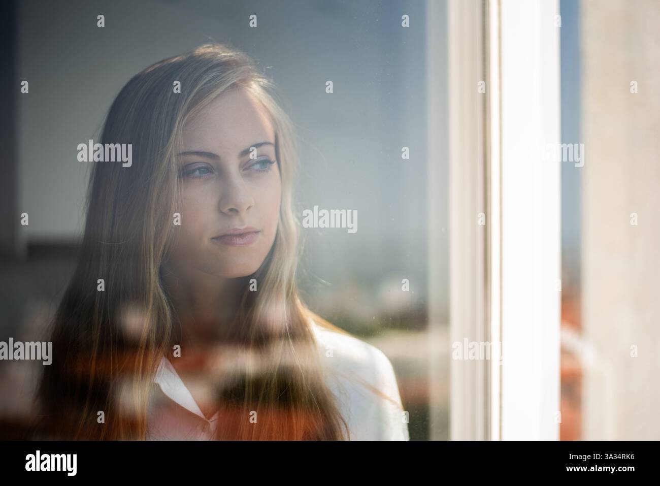 A thoughtful woman looks out of a window, her face softly reflected in ...