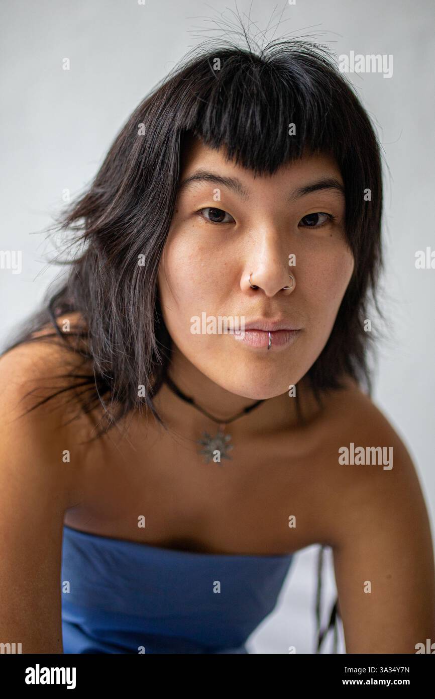 A close-up portrait of a young Asian woman with dark hair, bangs, and facial piercings. She ...