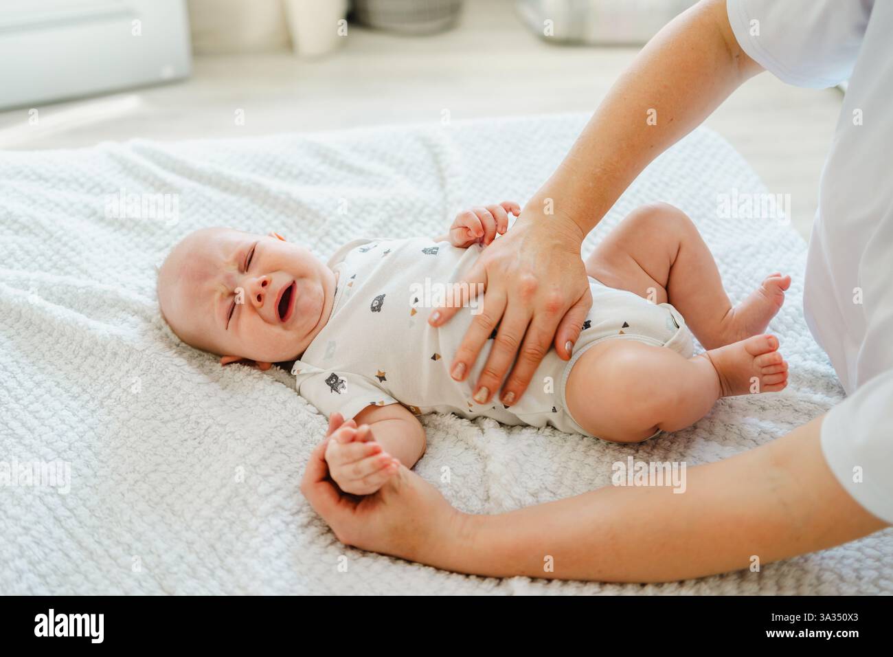 A mother comforts a crying baby lying on a soft white blanket. The ...