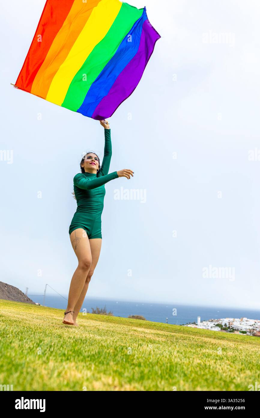 A joyful woman dances outdoors, waving a colorful LGBTQIA+ pride flag ...