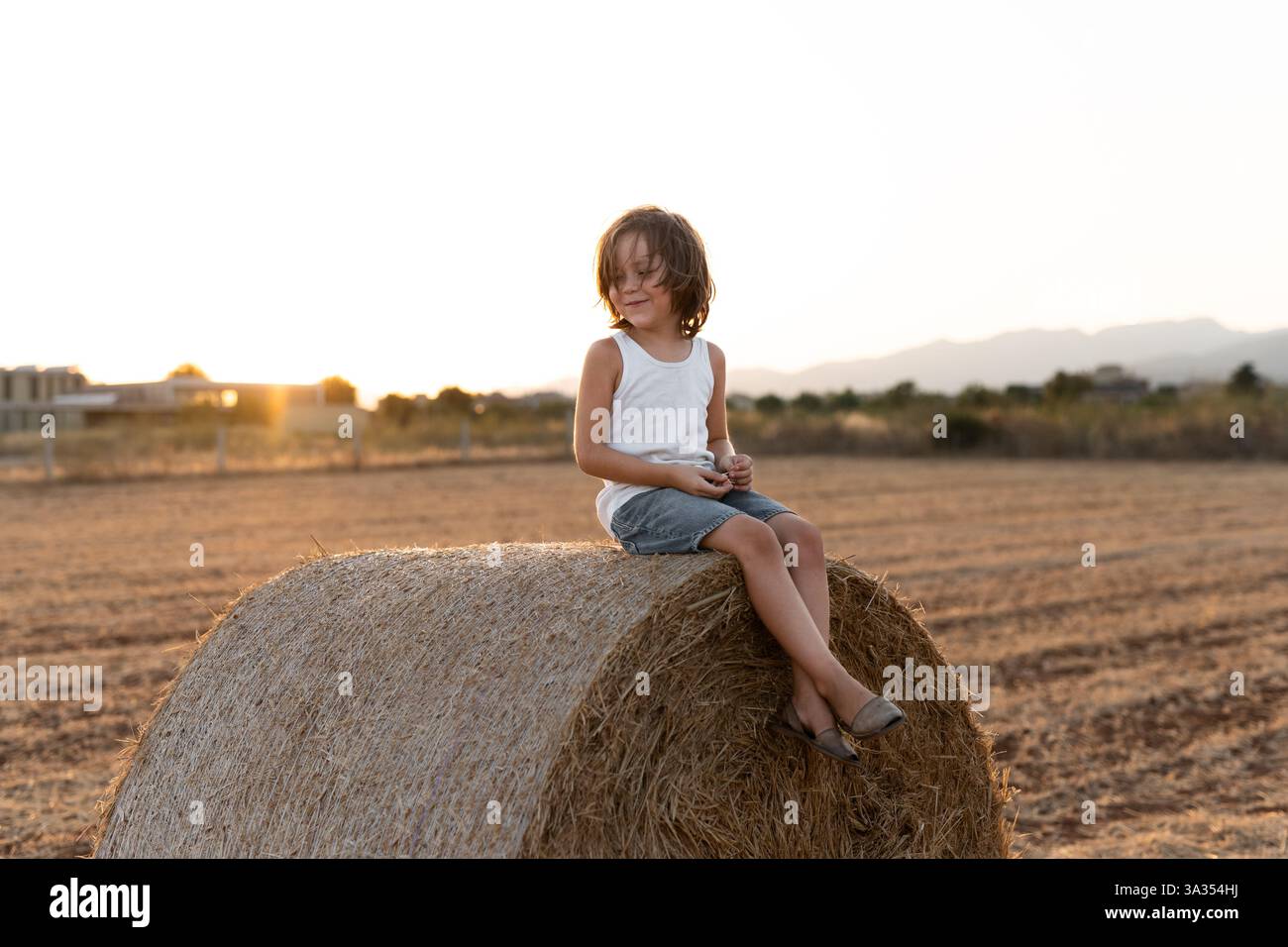A child in casual attire sits on a large hay bale in a field. The warm ...