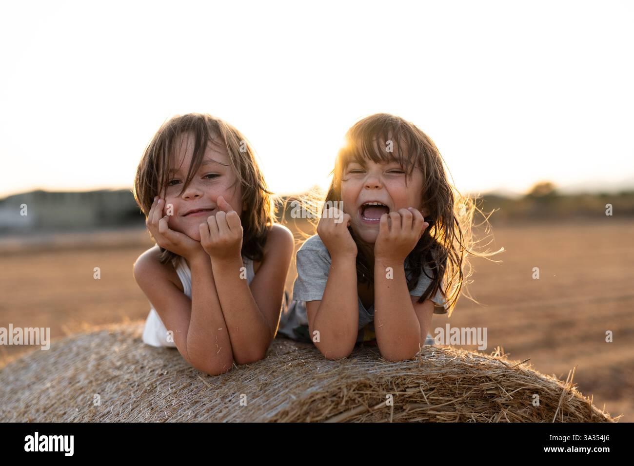 Two happy children lie on a hay bale, bathed in warm sunlight, laughing ...